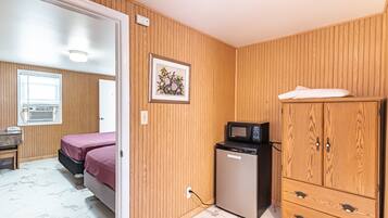 Quarto compacto com paredes de painel de madeira, cama com roupa de cama rosa, um pequeno frigorífico, micro-ondas e um armário com gavetas.