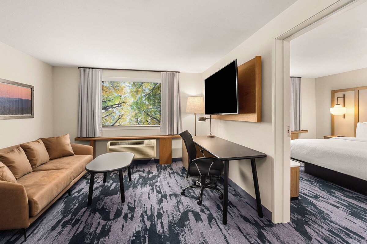 Quarto de hotel com sofá, escrivaninha, cadeira, televisão, cama e uma janela grande com cortinas.