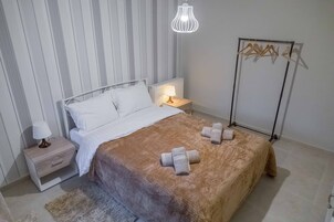 Uma cama bem arrumada com um cobertor castanho e duas toalhas, uma mesa de cabeceira com abajur, um radiador e um cabideiro.
