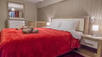 Cama bem arrumada com manta vermelha e toalhas dobradas, cabeceira de madeira, mesa de cabeceira com abajur e espelho sobre a cômoda.