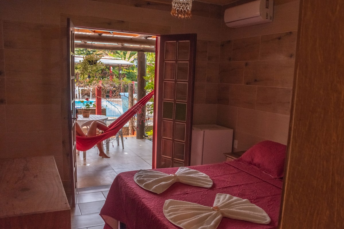 Quarto com cama, rede e vista para piscina e vegetação lá fora.