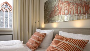 Quarto de hotel com uma janela grande, cama com roupa de cama branca e almofadas laranja, e um quadro pendurado na parede.