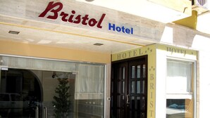 Exterior do Hotel Bristol, com entrada de vidro, plantas em vasos e uma placa sobre a porta.