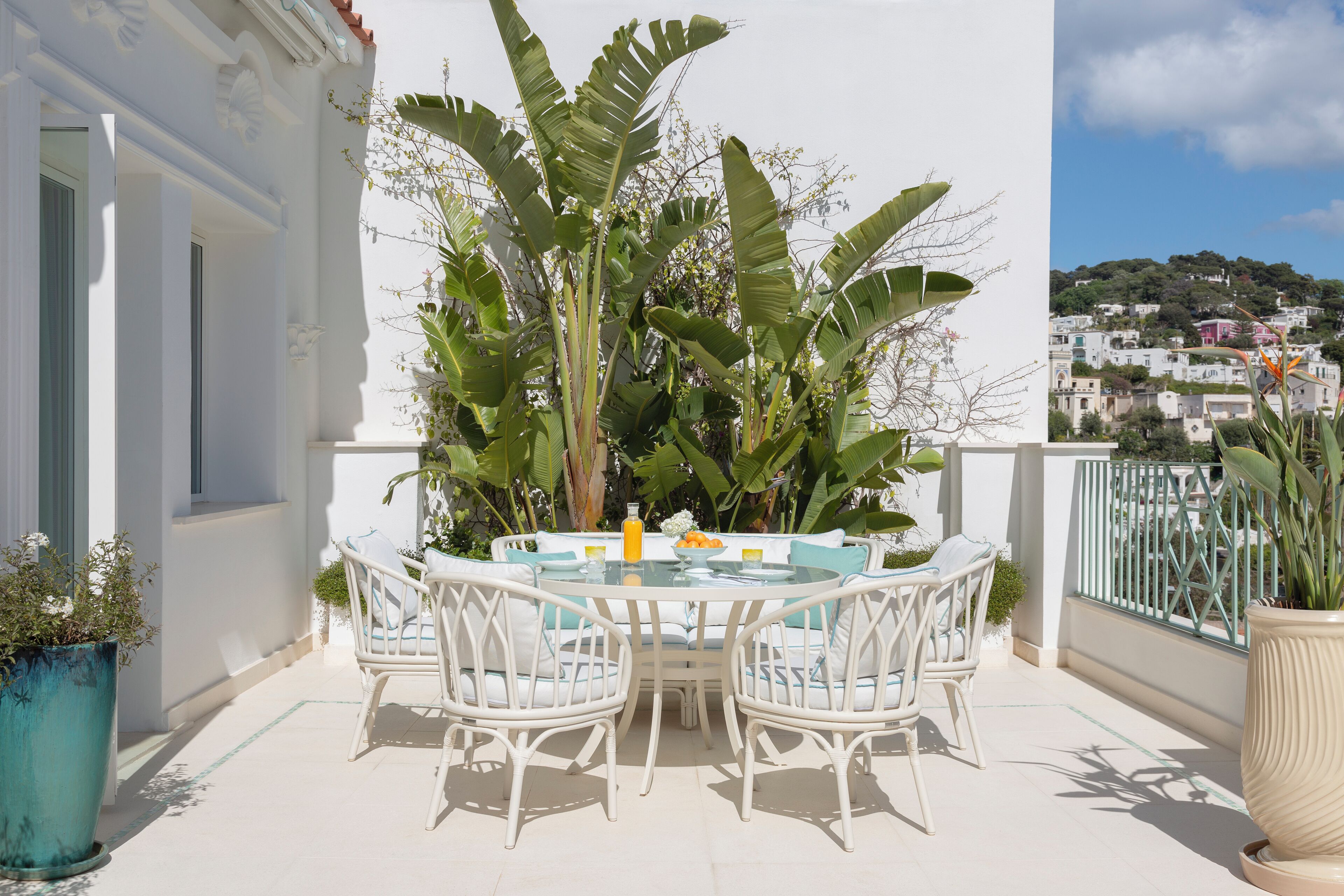 Mesa redonda com cadeiras brancas, rodeada por grandes plantas de bananeira, em uma varanda com guarda-corpo branco e vista para uma cidade no alto de uma colina.