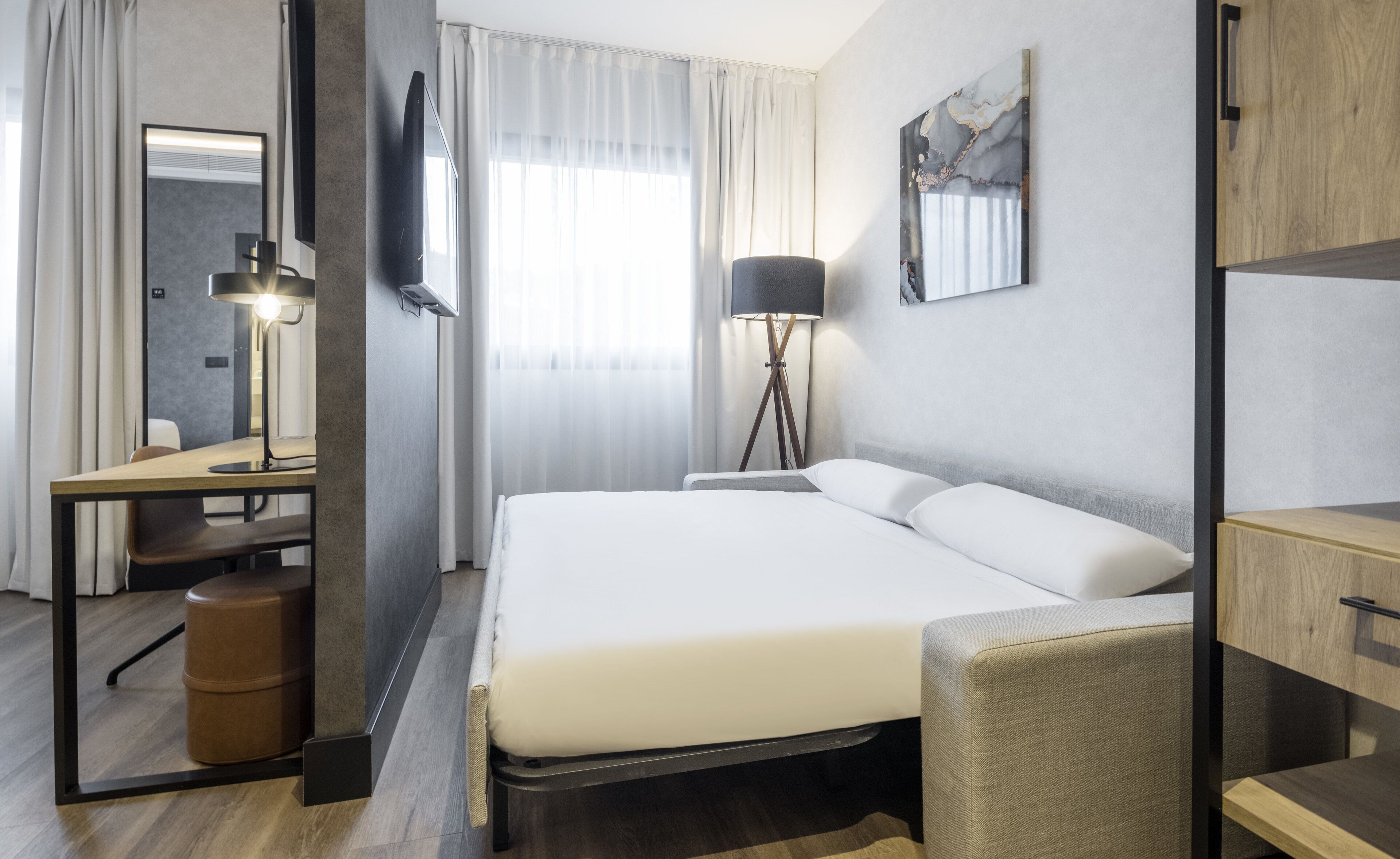 Quarto de hotel moderno com uma cama grande, uma mesa com cadeira, um abajur e uma TV de tela plana na parede.