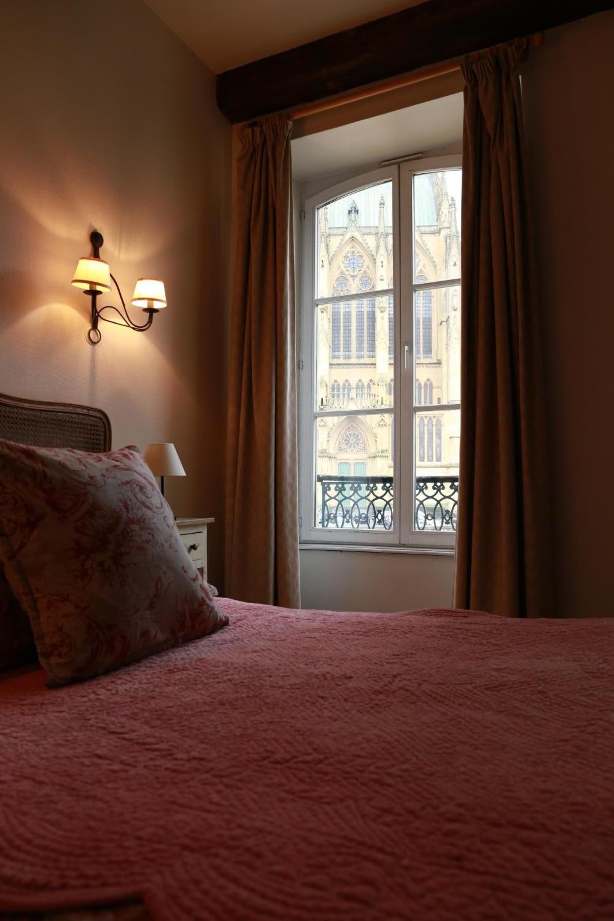 Quarto de hotel com uma janela ampla, uma cama com travesseiro floral e uma luminária fixada na parede.