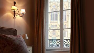 Quarto de hotel com uma janela ampla, uma cama com travesseiro floral e uma luminária fixada na parede.