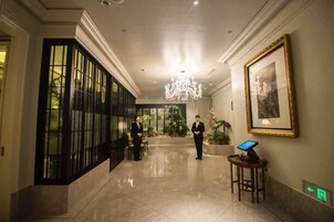 Um hall de hotel com um lustre, um balcão de recepção e uma grande janela com vista para o verde.