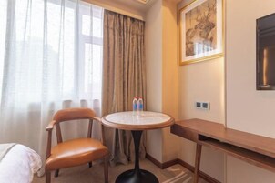 Quarto de hotel com cama, uma cadeira, uma mesa redonda com duas garrafas, um quadro e uma televisão na parede.