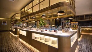 Uma área de buffet com um balcão central de serviço, diversas vitrines de vidro e iluminação suspensa.