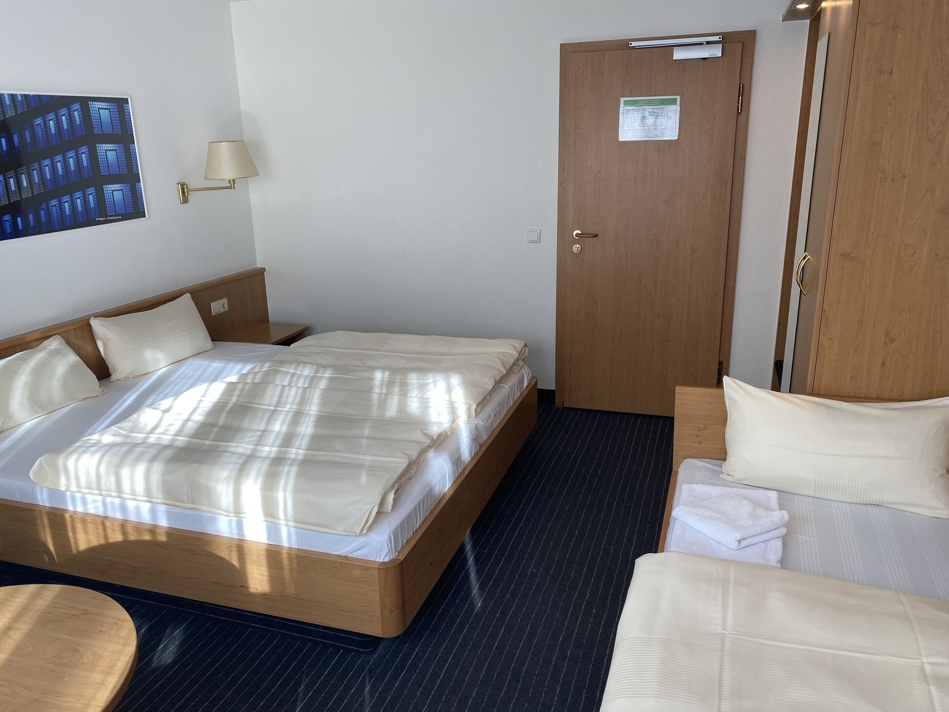 Quarto de hotel com uma cama grande, uma cama menor, um guarda-roupa de madeira, uma mesa de cabeceira, um abajur e um quadro pendurado na parede.