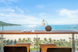 Um deck de madeira com vista para a praia e oceano, um par de cadeiras e uma mesa com um livro e um objeto decorativo.
