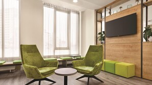 Uma área de espera moderna com duas cadeiras verdes, uma mesa redonda pequena e uma televisão de tela plana grande fixada em uma parede de madeira.