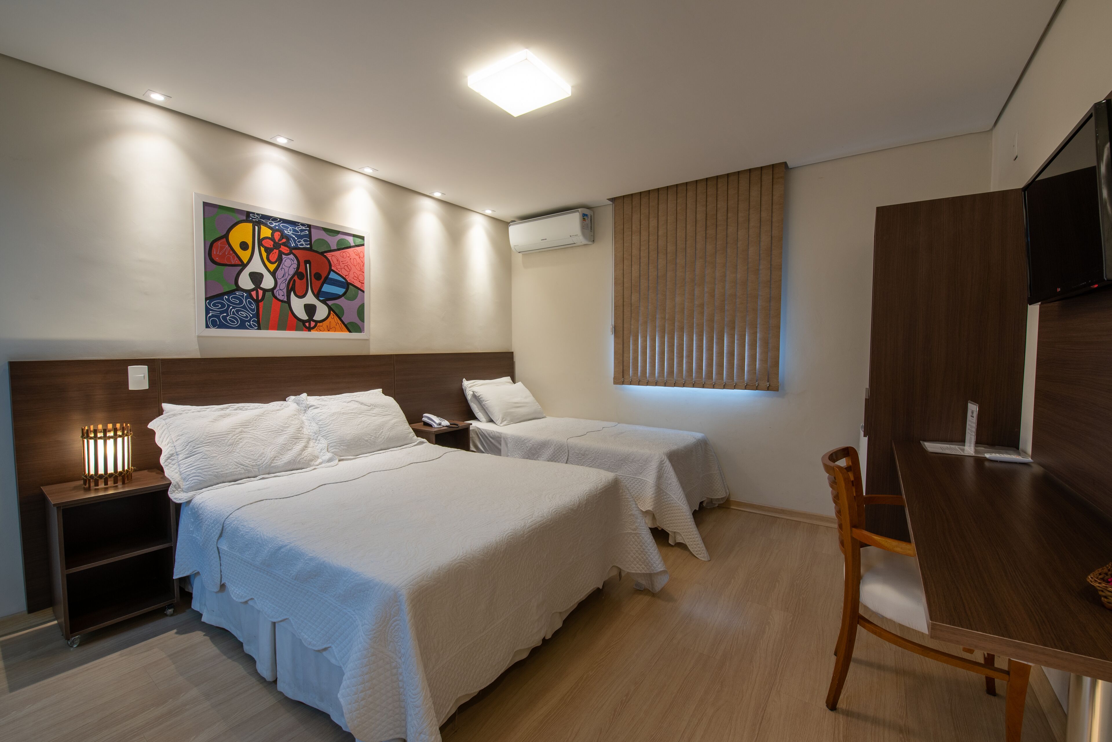 Quarto de hotel com duas camas, uma escrivaninha, televisão, ar condicionado, um quadro e janela com persianas.