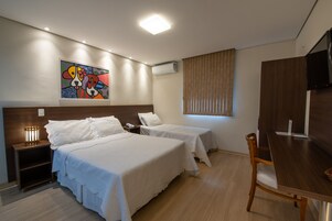 Quarto de hotel com duas camas, uma escrivaninha, televisão, ar condicionado, um quadro e janela com persianas.