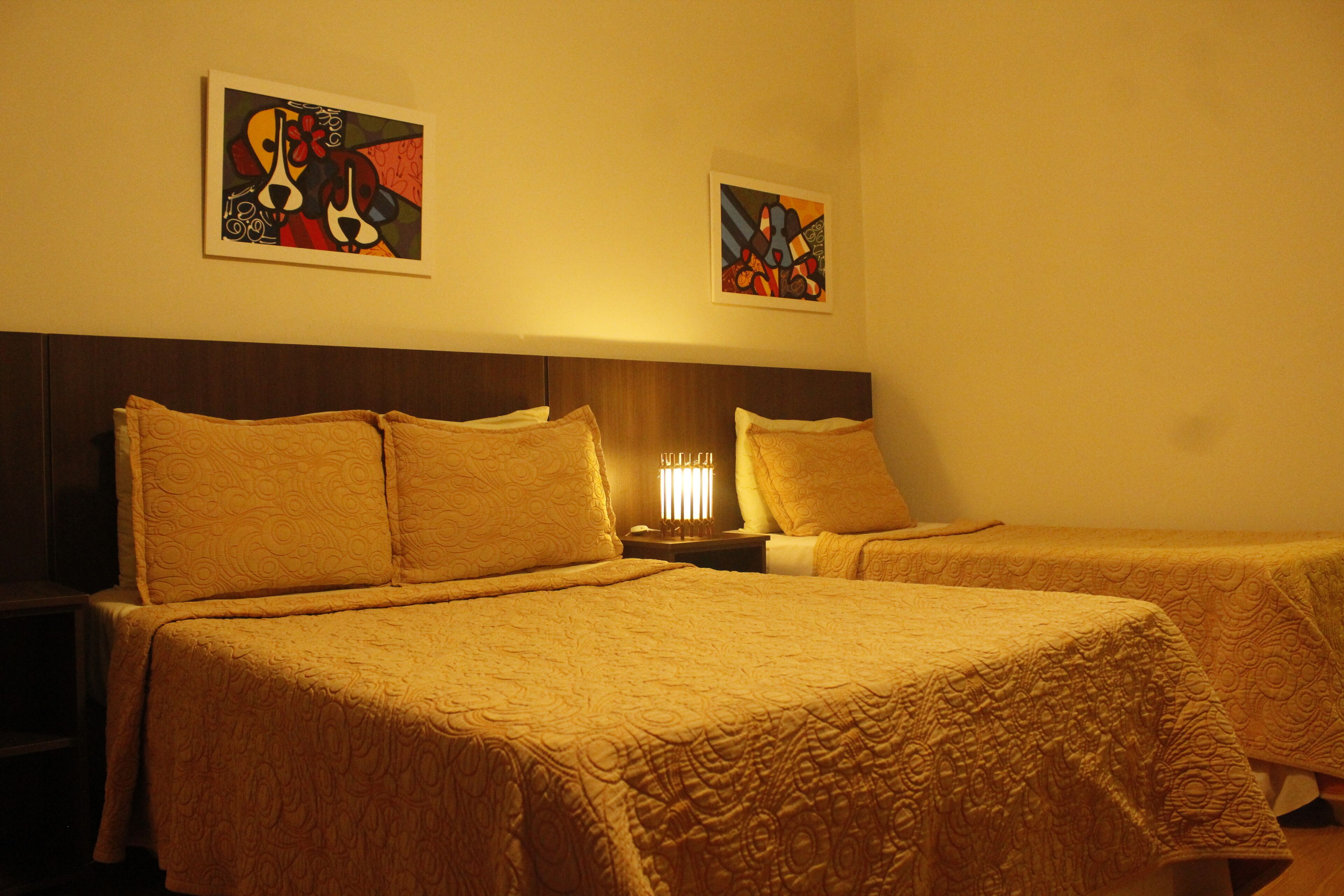 Quarto de hotel com duas camas, mesa de cabeceira com abajur e duas pinturas abstratas na parede.