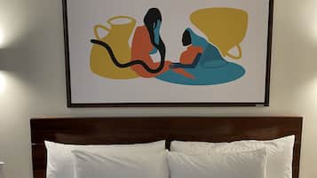 Quadro abstrato emoldurado numa parede acima de uma cama com almofadas brancas e cabeceira de madeira.