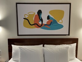 Quadro abstrato emoldurado numa parede acima de uma cama com almofadas brancas e cabeceira de madeira.