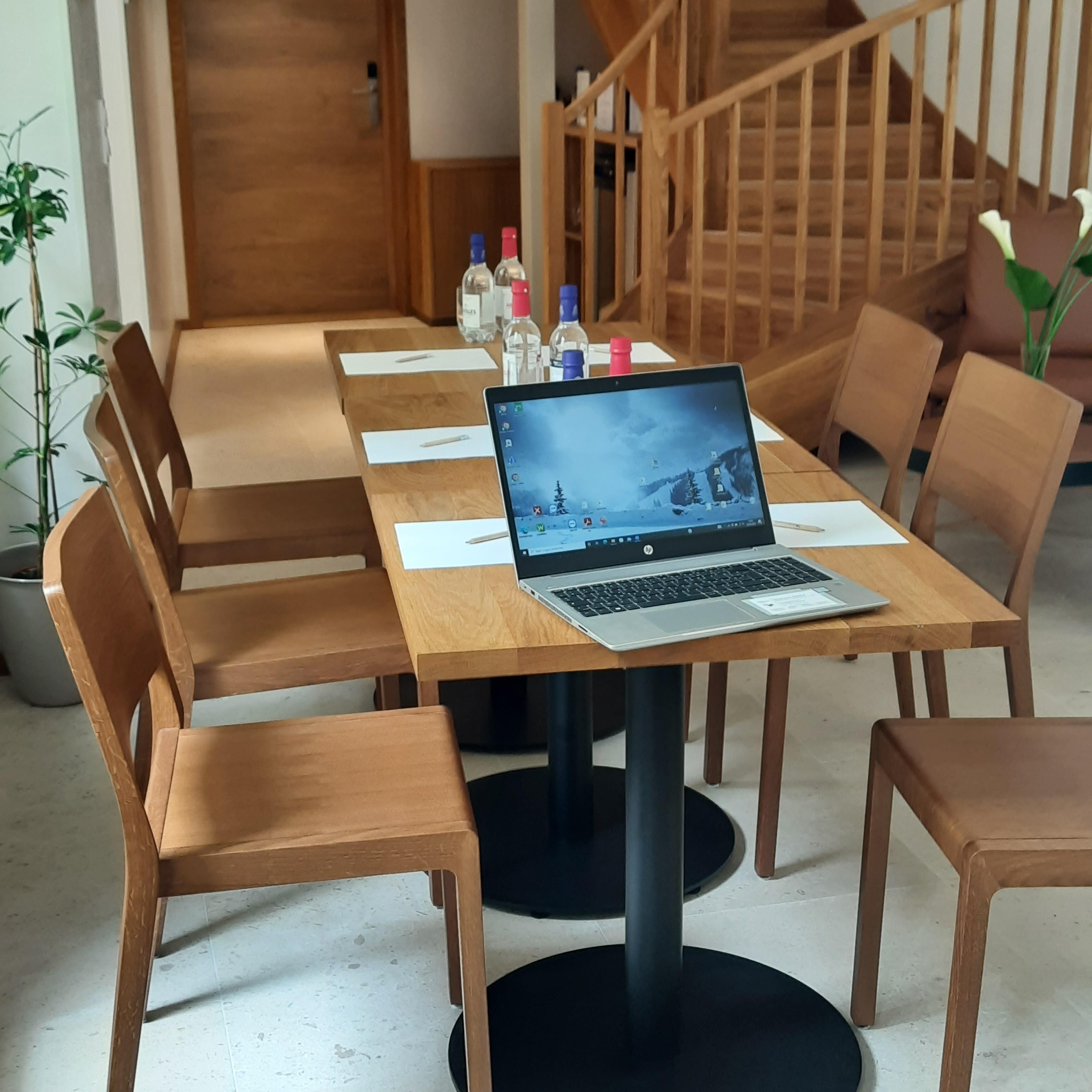 Uma mesa de jantar de madeira com um laptop, papéis e canetas. Cadeiras circundam a mesa, e uma escadaria é visível ao fundo.