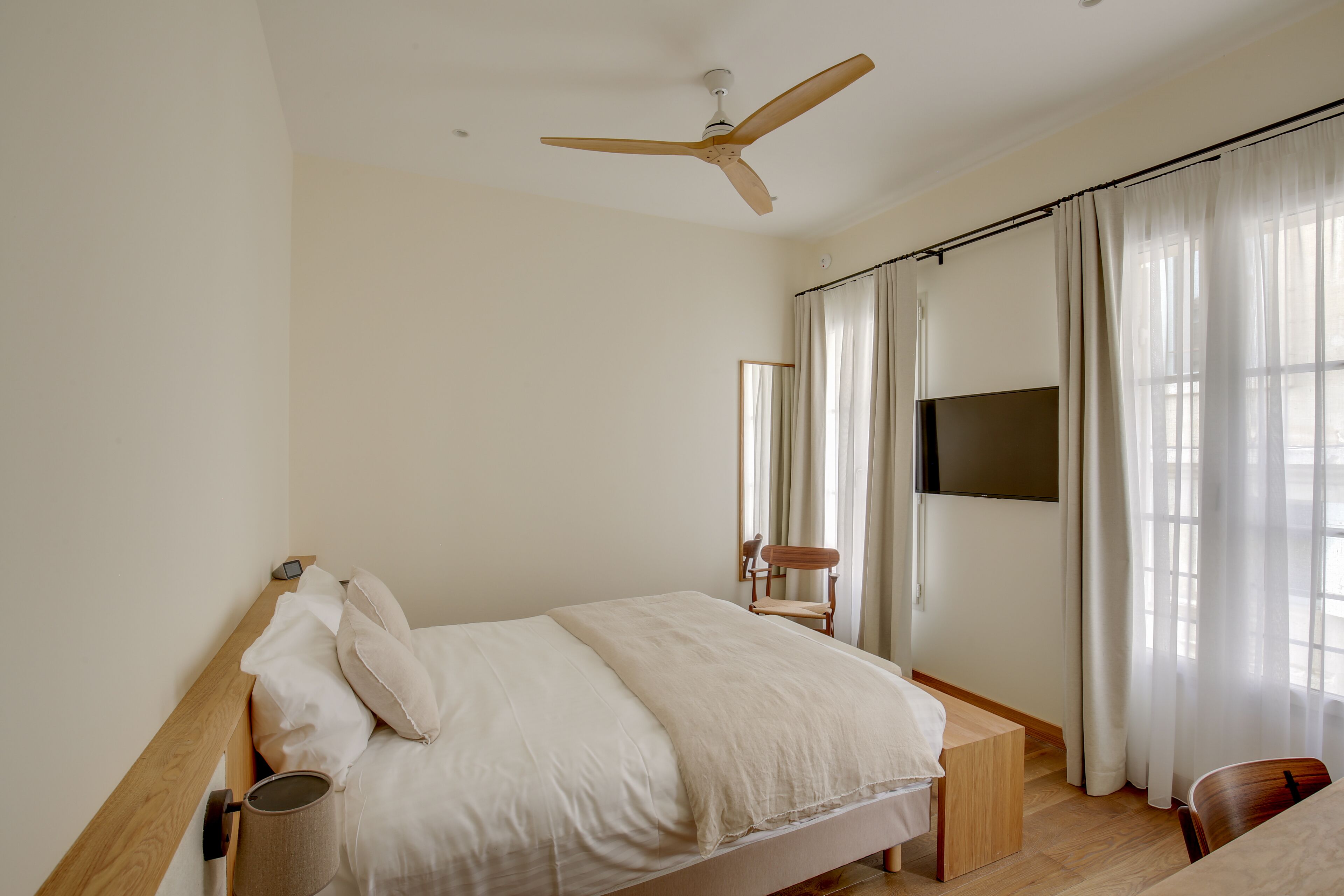 Quarto com cama, ventilador de teto, televisão na parede e janela com cortinas.