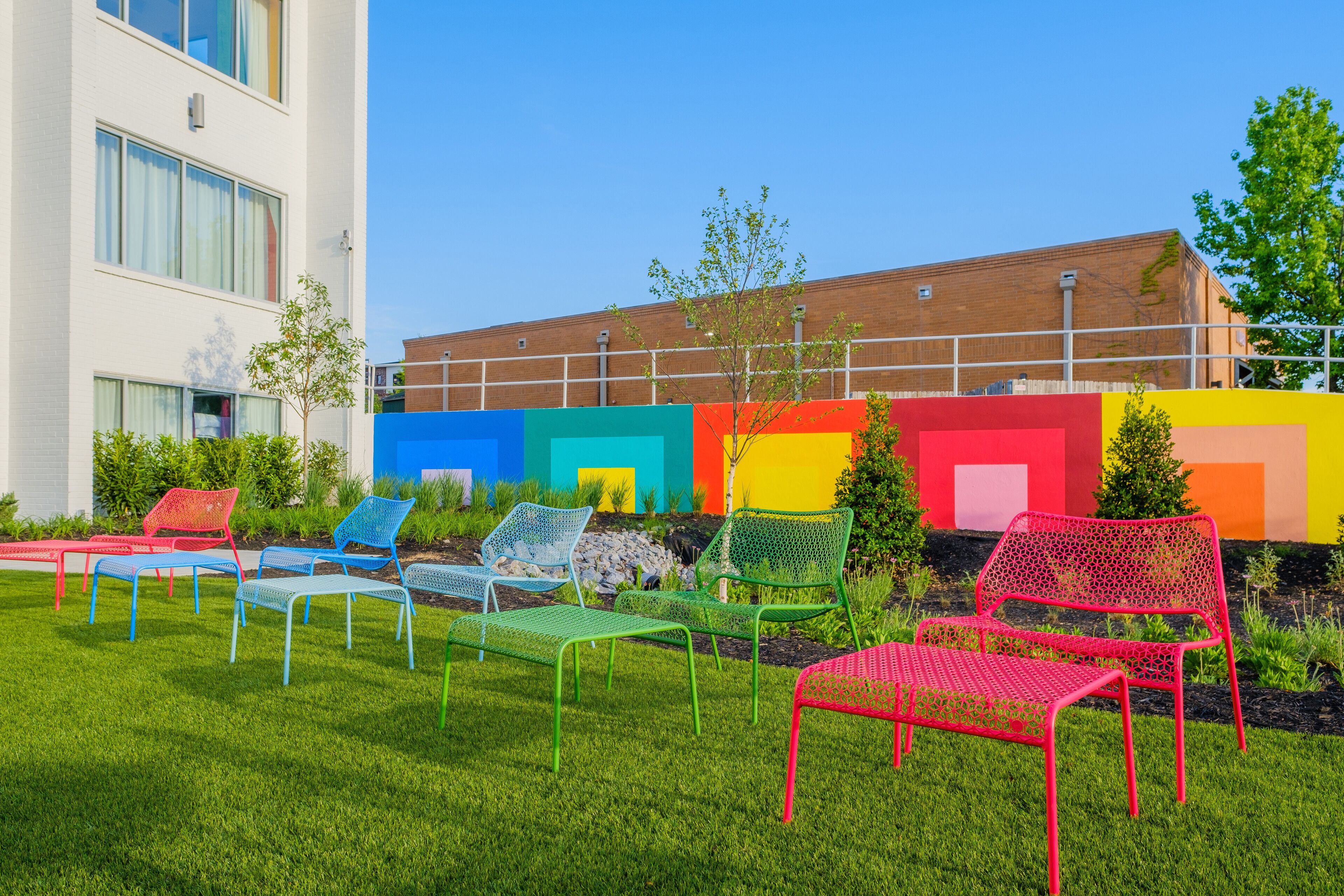 Uma área externa gramada com cadeiras de metal coloridas e um edifício com fachada multicolorida ao fundo.