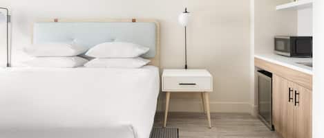 Quarto bem arrumado com cama, mesa de cabeceira, abajur e um micro-ondas numa prateleira.
