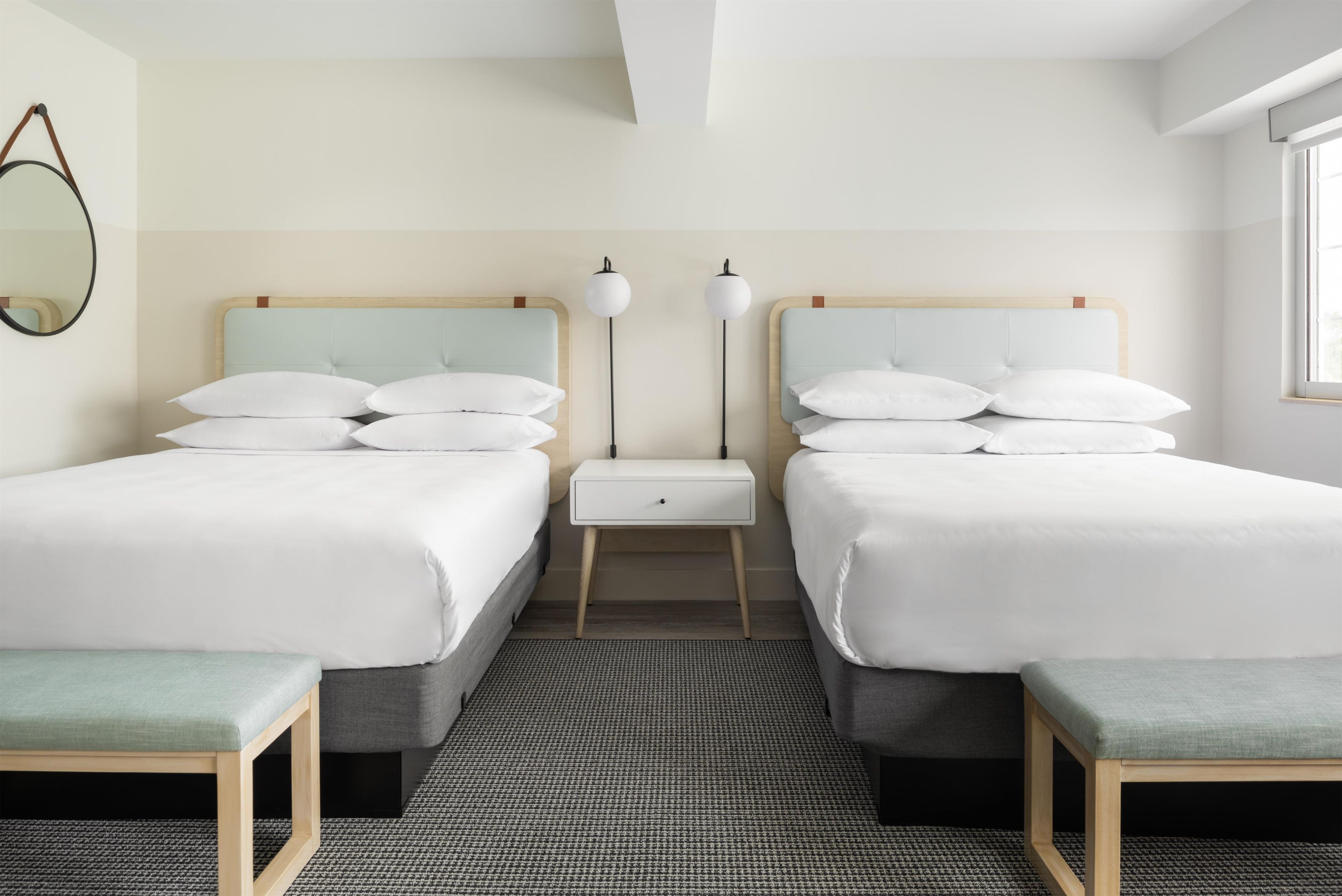 Quarto de hotel com duas camas, uma mesa de cabeceira, um abajur e um espelho na parede.