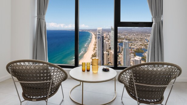 Quarto com uma grande janela com vista para a praia e para a cidade, com duas cadeiras de vime e uma mesa pequena com um jarro e copos.