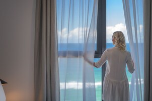 Uma pessoa em pé perto de uma janela com cortinas finas, apreciando a vista do oceano.
