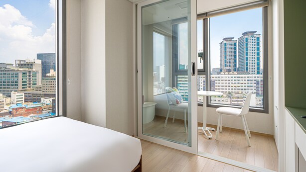 Quarto de hotel moderno com uma cama grande, uma varanda com porta de vidro, uma pequena mesa e uma cadeira branca.