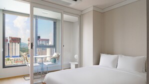 Quarto de hotel com uma cama grande, uma mesa pequena, uma cadeira e vista da cidade através de uma porta de vidro deslizante.