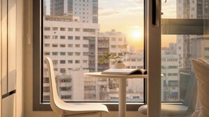Quarto moderno com vista para a cidade ao pôr do sol, uma cadeira branca, uma pequena mesa com um livro e uma planta, e uma cama com roupa de cama branca.