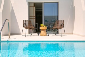 Uma área à beira da piscina com duas cadeiras de vime, uma pequena mesa com bebidas, a própria piscina e a entrada de um edifício.