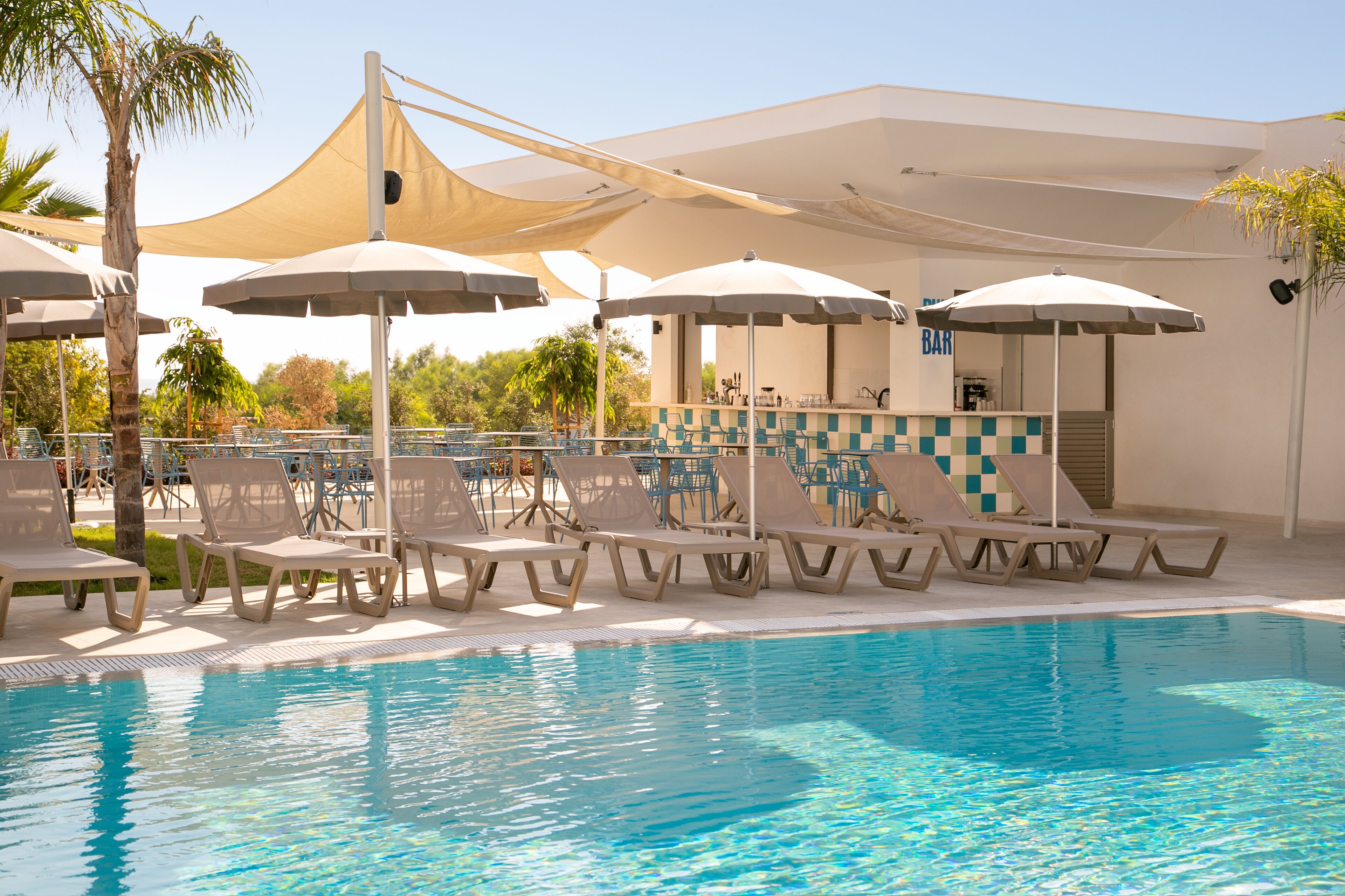 Área da piscina com espreguiçadeiras e guarda-sóis, uma piscina e um bar com parede de azulejos xadrez.