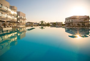 Hotel com piscina refletindo edifícios e palmeiras, pôr do sol ao fundo.