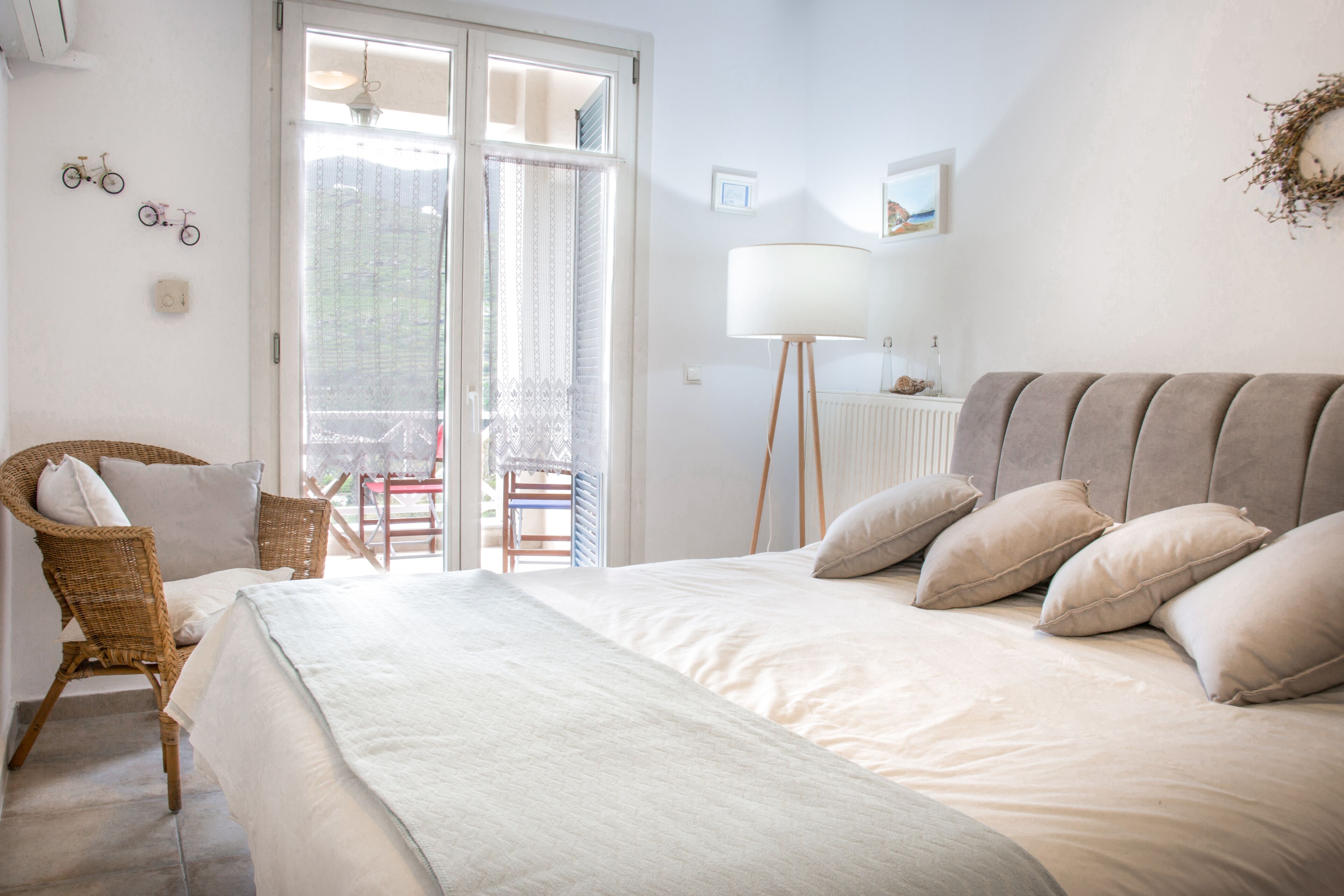 Quarto com cama, uma cadeira de vime, luminária de chão e vista através de uma porta de vidro.