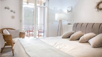 Quarto com cama, uma cadeira de vime, luminária de chão e vista através de uma porta de vidro.