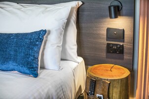 Cama bem arrumada com roupa de cama branca e azul, luminária fixada na parede, tomada e toco de madeira com controle remoto.
