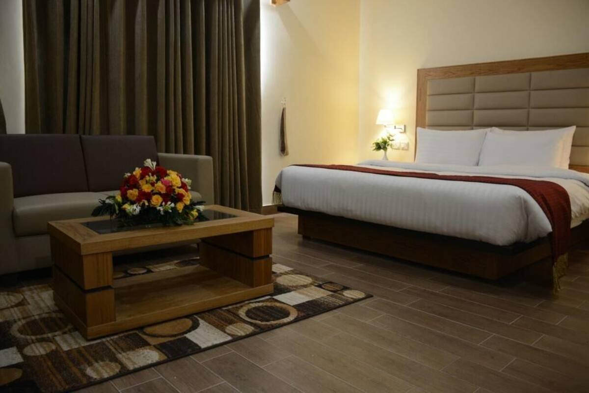 Quarto de hotel com uma cama, um sofá, uma mesa de centro de madeira e um arranjo floral, e um tapete estampado.