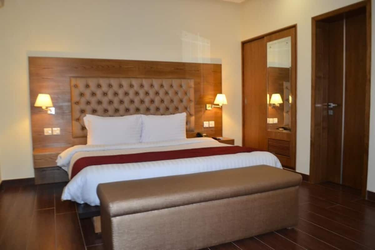 Quarto de hotel com uma cama grande, duas luminárias de cabeceira, uma cabeceira de madeira, um banco e um guarda-roupa.