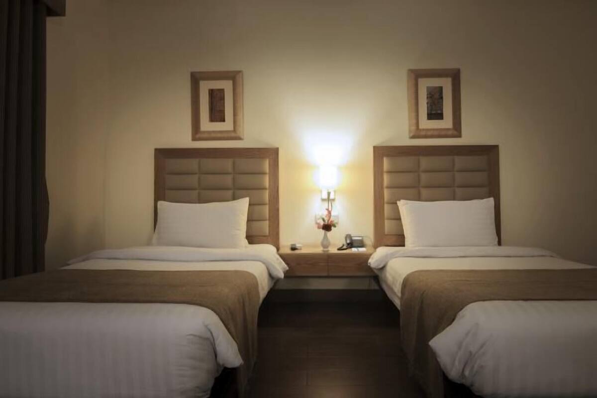 Quarto de hotel com duas camas, mesa de cabeceira com abajur e quadros nas paredes.