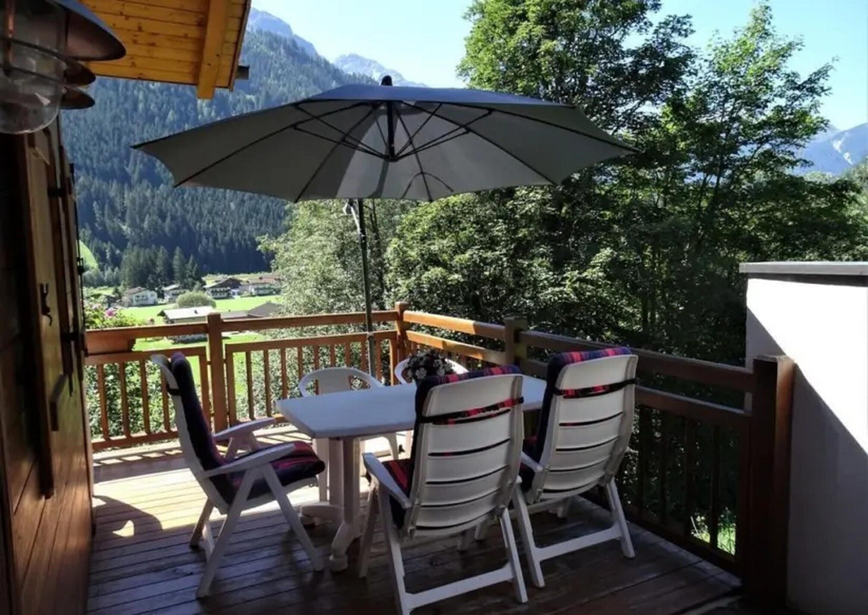 Um deck de madeira com mesa, cadeiras e um guarda-sol, com vista para uma paisagem montanhosa.