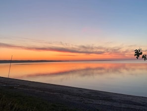 Um lago calmo ao pôr do sol, com céu claro, um píer à esquerda e uma árvore com folhagem à direita.