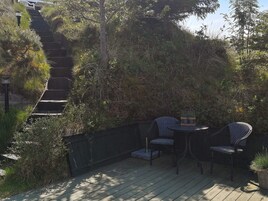 Um deck de madeira com duas cadeiras e uma pequena mesa, cercado por folhagem densa e uma escada que sobe.