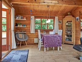 Interior de uma cabana de madeira com mesa de jantar, cadeiras, pequena cozinha e janela com vista para o verde.