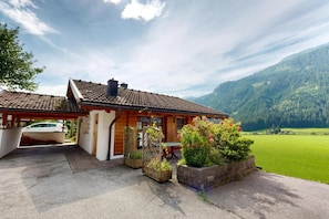 Uma casa de madeira com telhado de telhas, um carro estacionado na entrada e uma vista de um vale verde e montanhas ao fundo.