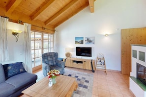 Uma sala de estar acolhedora com um sofá, uma poltrona xadrez, uma mesa de centro com flores, uma televisão e uma lareira.