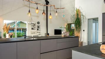 Cozinha moderna com ilha central, iluminação pendente e vista para uma casa verde.