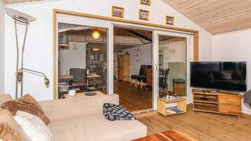 Sala de estar com sofá, televisão e mesa de madeira. Uma porta de vidro deslizante dá acesso a uma área de jantar.