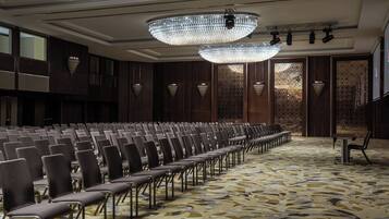 Sala de conferências com fileiras de cadeiras, um tapete estampado e lustres.